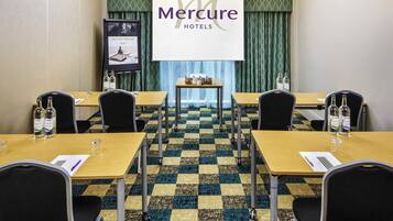 Sala de conferências com carpete xadrez, quatro mesas, cadeiras, tela de projeção com a marca 'Mercure Hotels' e garrafas nas mesas.