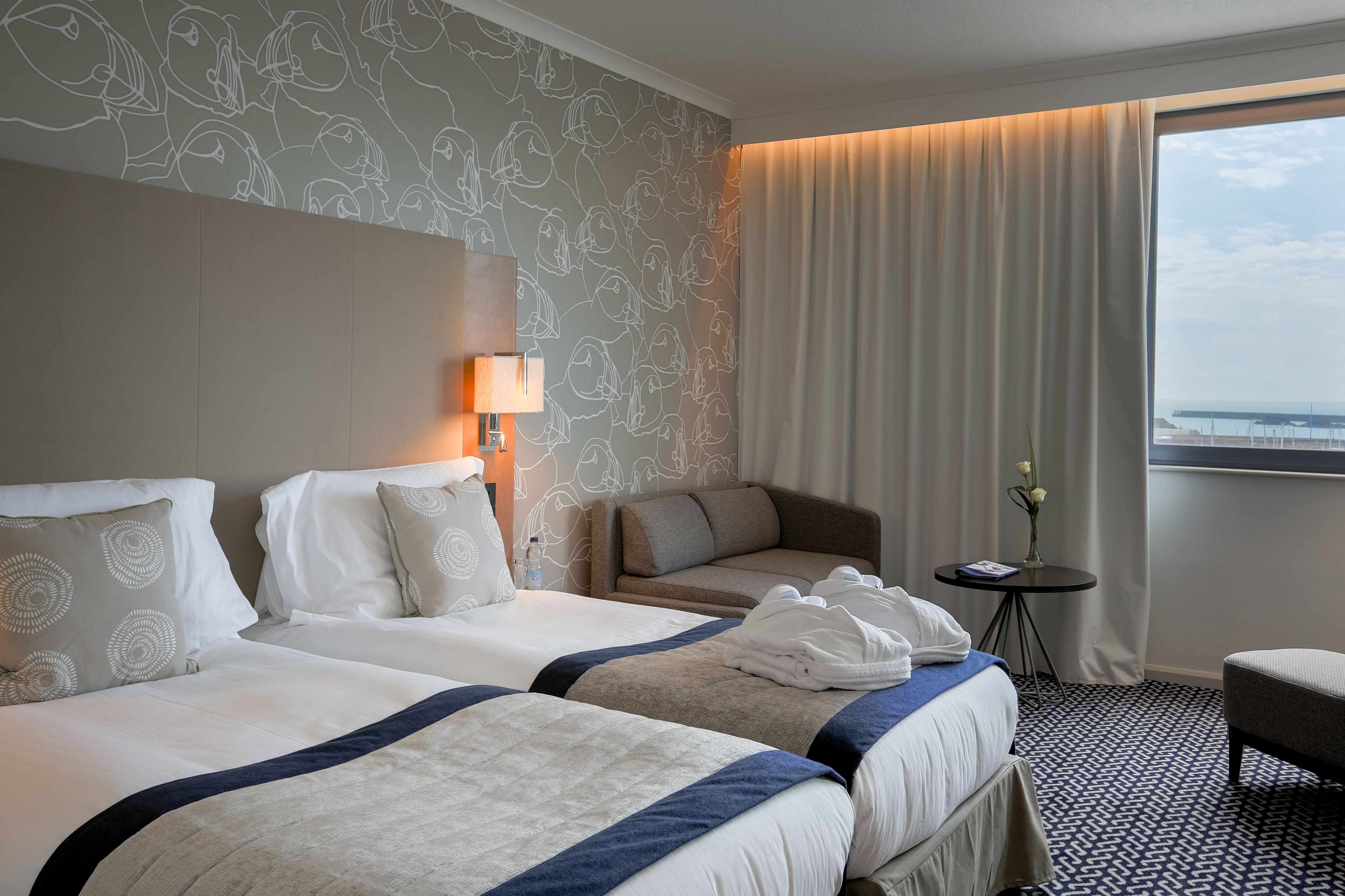 Quarto de hotel com duas camas, um sofá, uma mesinha com um vaso e vista para o mar.