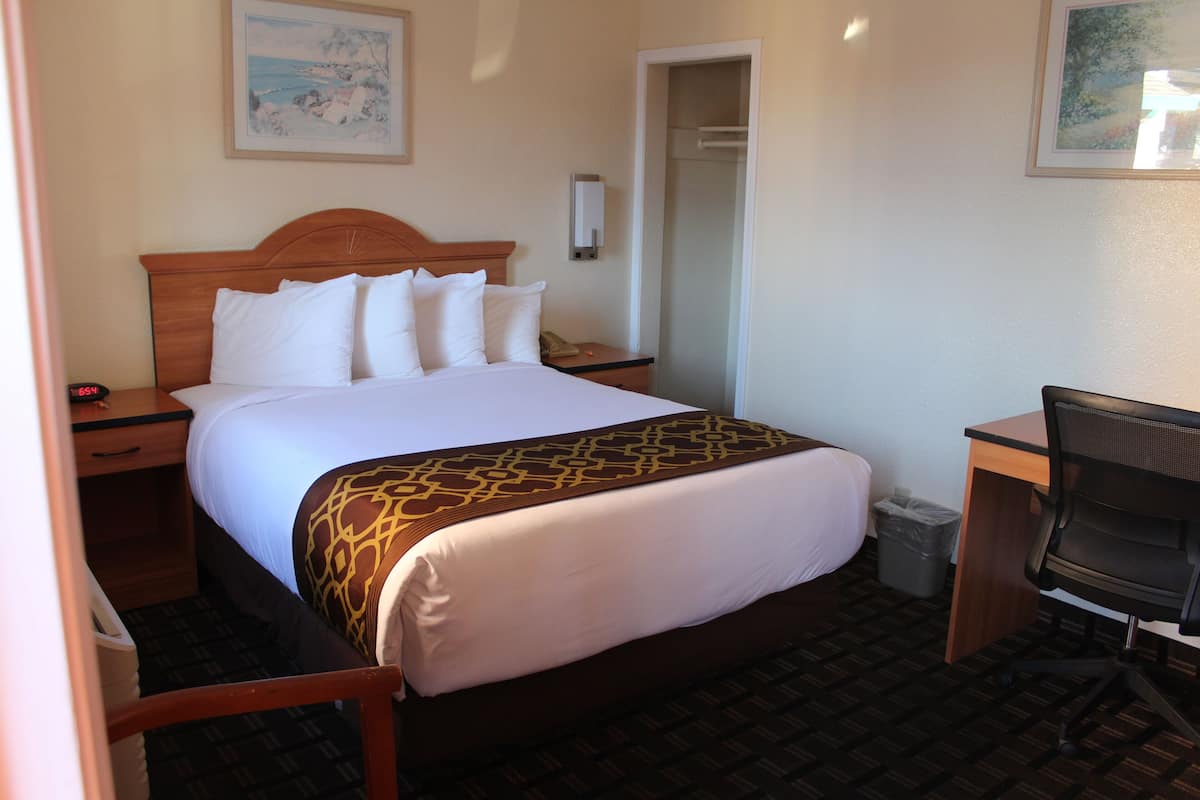 Quarto de hotel com uma cama grande, uma escrivaninha e uma cadeira. Há uma porta que dá para outro cômodo e uma lixeira.