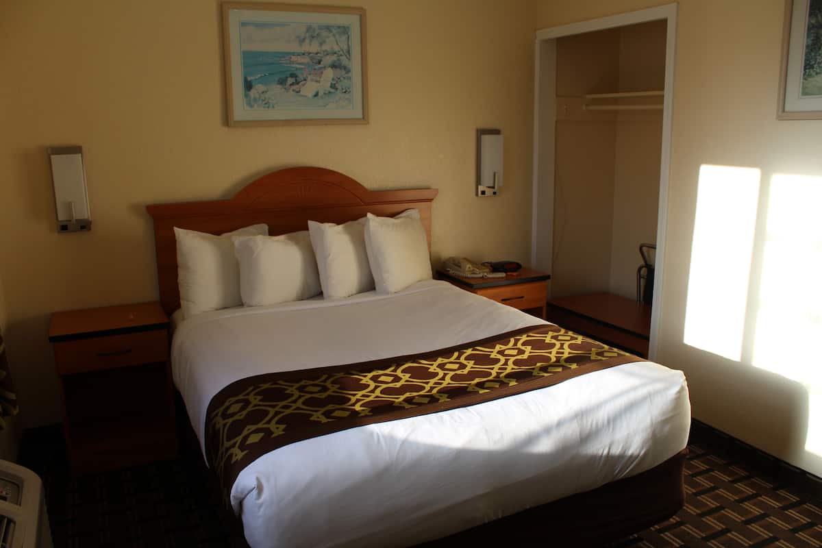 Quarto de hotel com uma cama grande, cabeceira de madeira, mesa de cabeceira, espelho e janela com luz natural.