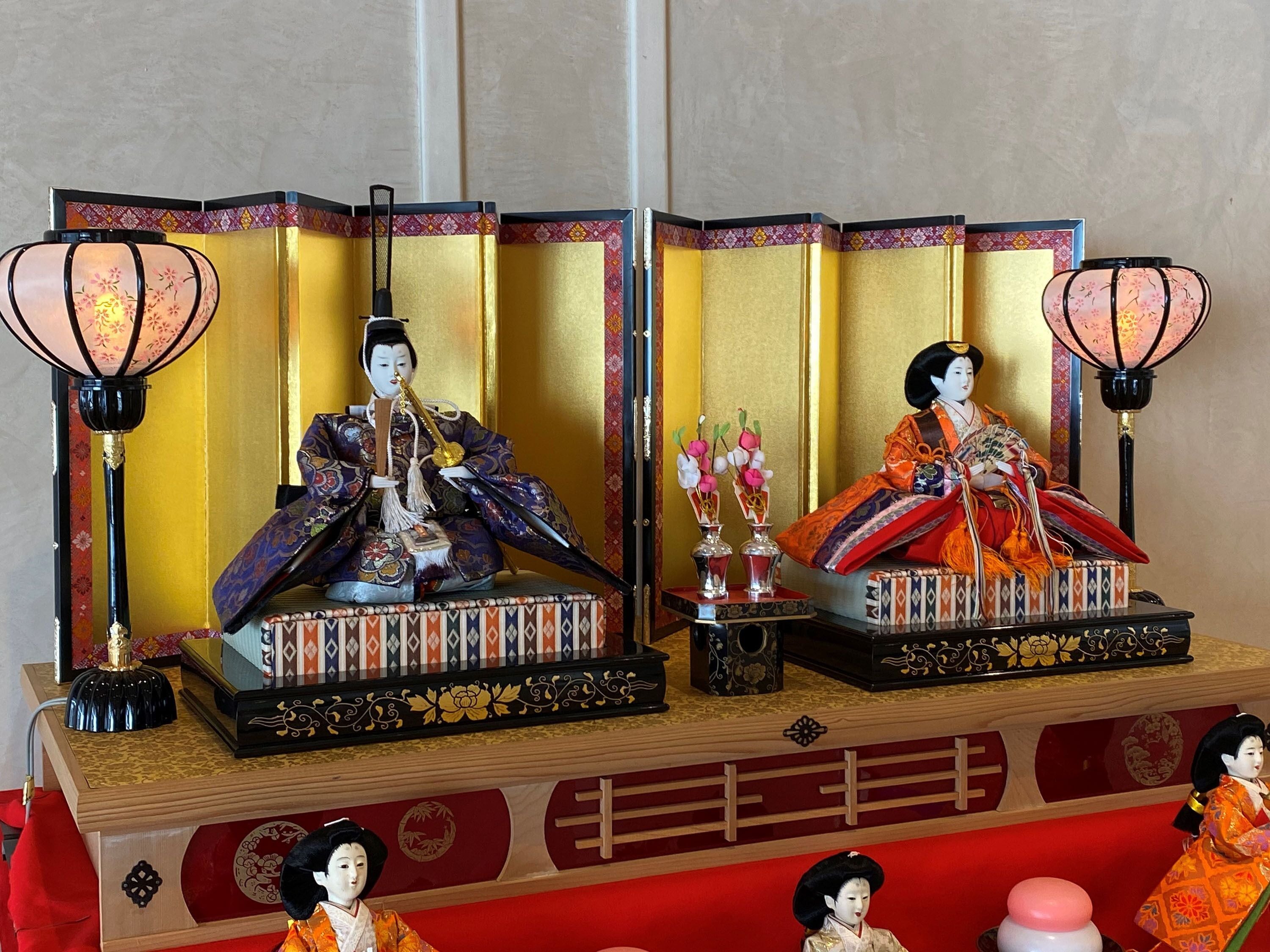 Duas bonecas japonesas tradicionais sobre uma plataforma de madeira, com um fundo vermelho e dourado, e acompanhadas por duas lâmpadas pretas.