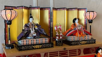 Duas bonecas japonesas tradicionais sobre uma plataforma de madeira, com um fundo vermelho e dourado, e acompanhadas por duas lâmpadas pretas.
