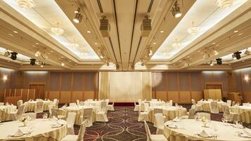 Uma sala de banquetes com mesas redondas preparadas para refeições, equipada com lustres e iluminação embutida.