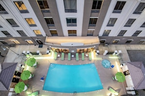 Área de piscina no terraço com espreguiçadeiras e guarda-sóis, cercada por um edifício alto com diversas janelas.