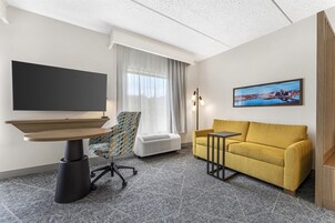Quarto de hotel com televisão de tela plana, escrivaninha com cadeira, sofá, mesa pequena, abajur e um quadro na parede.