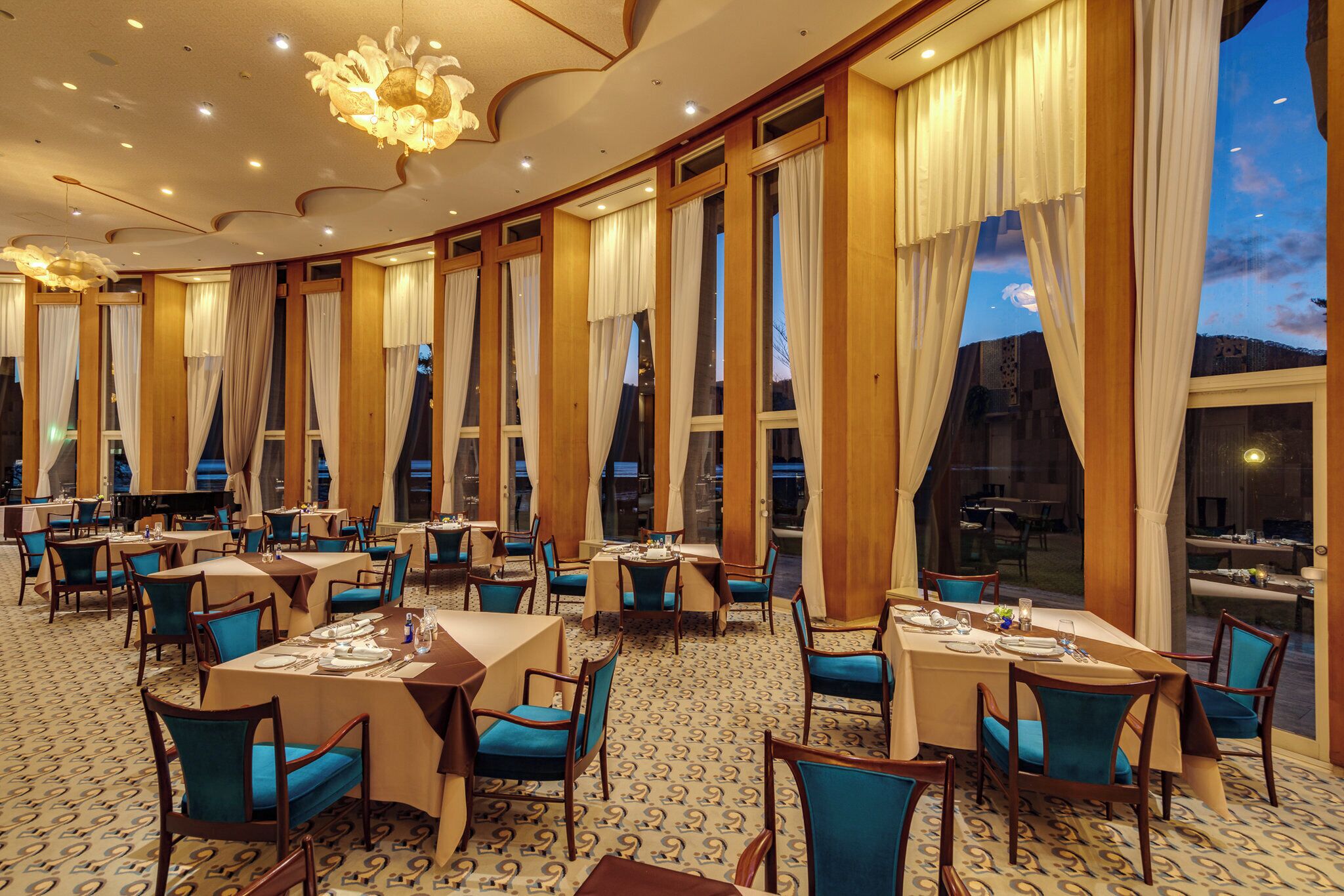 Uma sala de jantar de hotel com mesas preparadas para hóspedes, grandes janelas com cortinas e um tapete estampado.