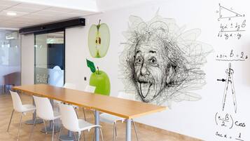Uma sala de conferências com uma mesa comprida, cadeiras brancas e um mural na parede com equações matemáticas e uma maçã verde.