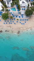 Vista aérea de um resort à beira-mar, com piscina, vários guarda-sóis azuis e um mar azul-turquesa cristalino.