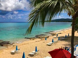 Uma praia com águas turquesa cristalinas, areal arenoso e cadeiras de praia com guarda-sóis azuis.