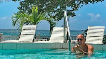 Área à beira da piscina com espreguiçadeiras, vista desobstruída do oceano e uma árvore oferecendo sombra.