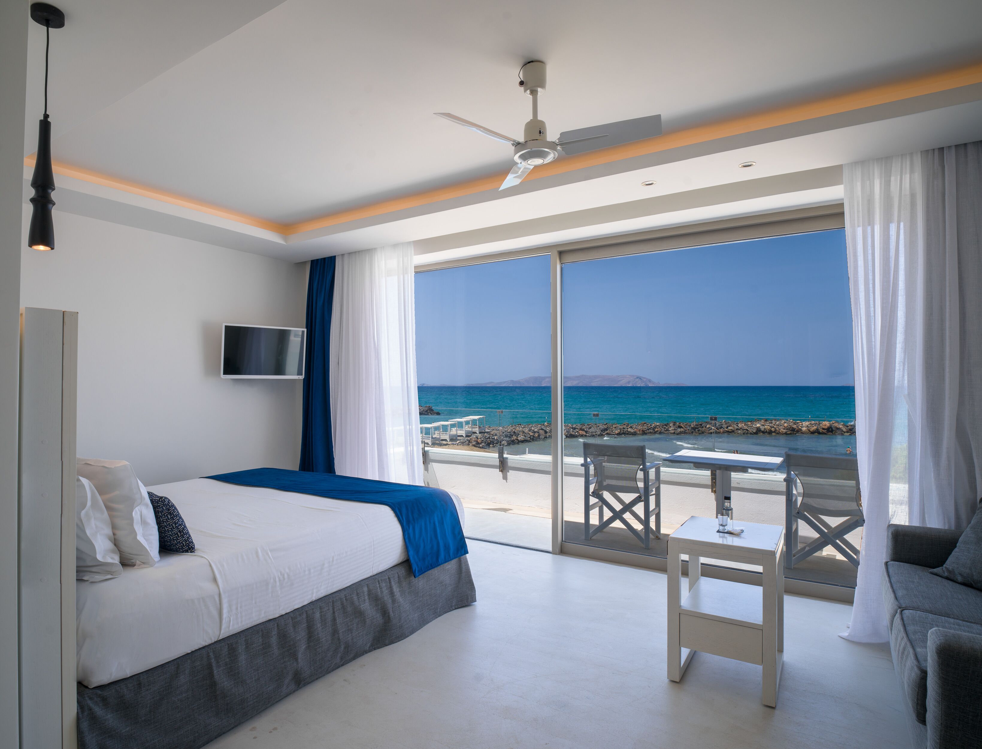 Quarto com cama, TV, ventilador de teto e vista para o mar através de grandes janelas.