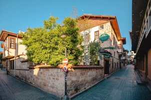 Uma rua de paralelepípedos estreita, ladeada por edifícios de pedra tradicionais, uma árvore com frutos maduros e uma placa indicando um restaurante local.