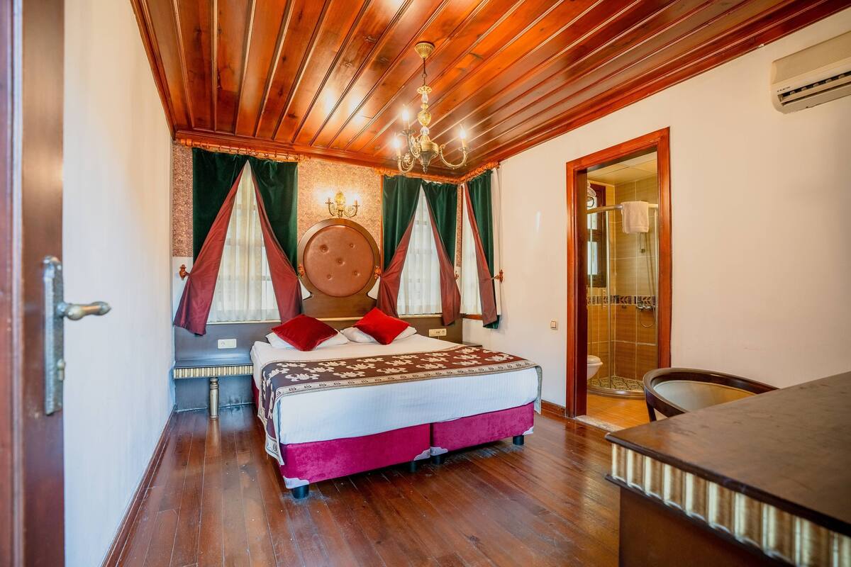 Quarto com teto de madeira, cama com cabeceira, escrivaninha e área de chuveiro.