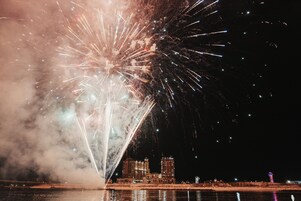 Vista noturna de uma orla com show de fogos de artifício, prédios e um píer.