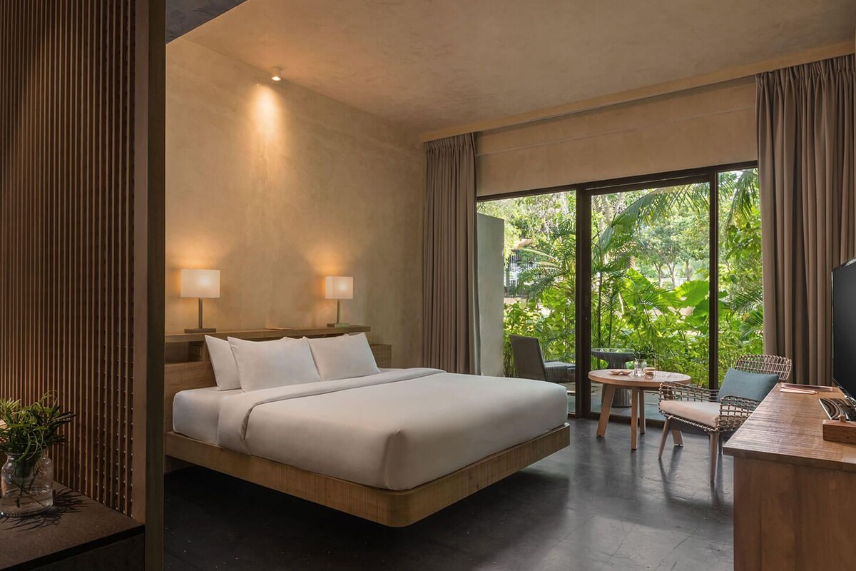 Quarto de hotel com cama, mesas de cabeceira, escrivaninha, cadeira, televisão e uma grande janela com vista para o verde.