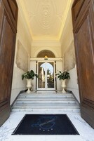 Uma entrada imponente com degraus de mármore, um tapete preto com o brasão do hotel e duas grandes plantas em vasos.