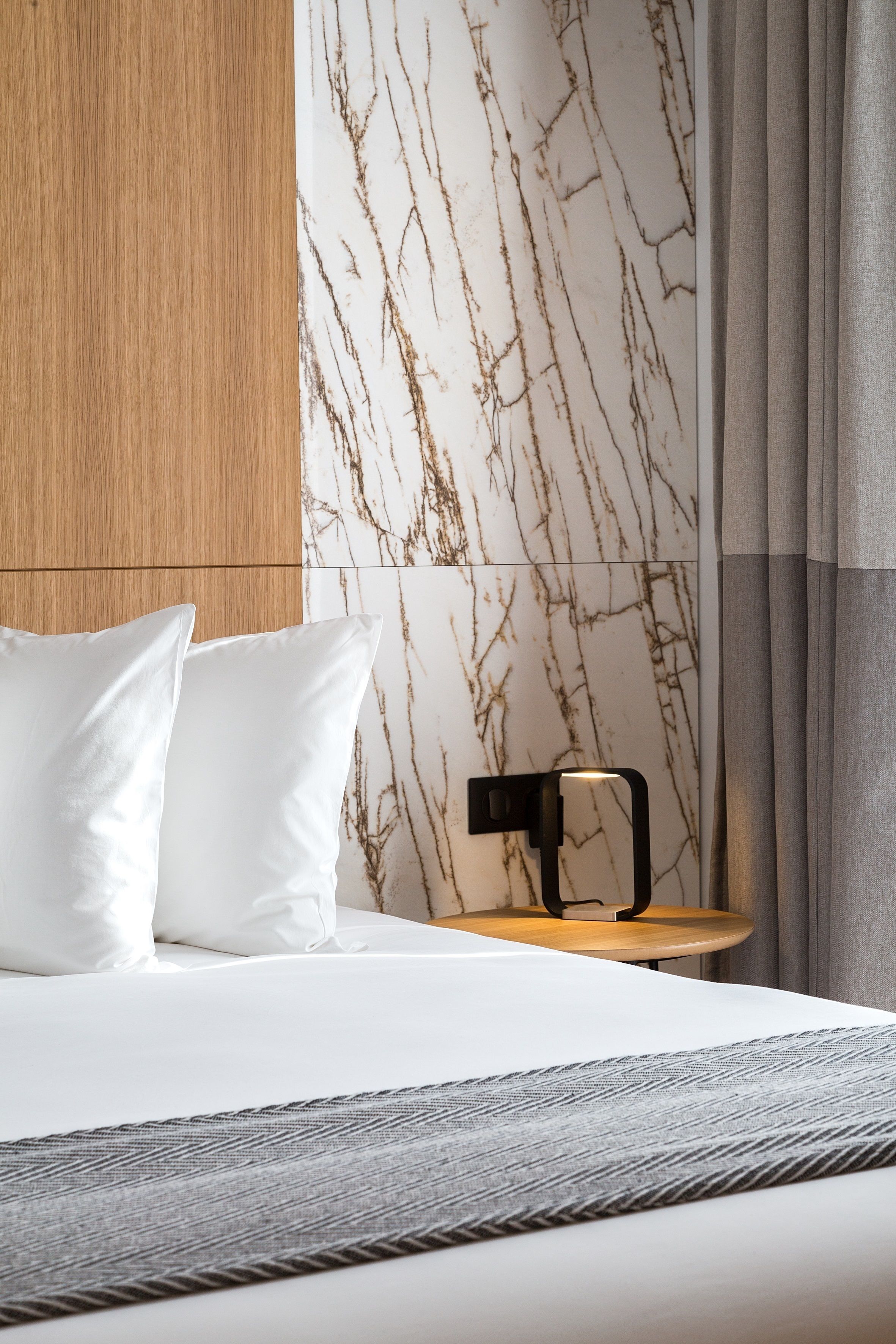 Uma cama bem arrumada com roupa de cama branca, uma mesa de cabeceira de madeira com uma luminária moderna e uma parede com padrão que lembra mármore.
