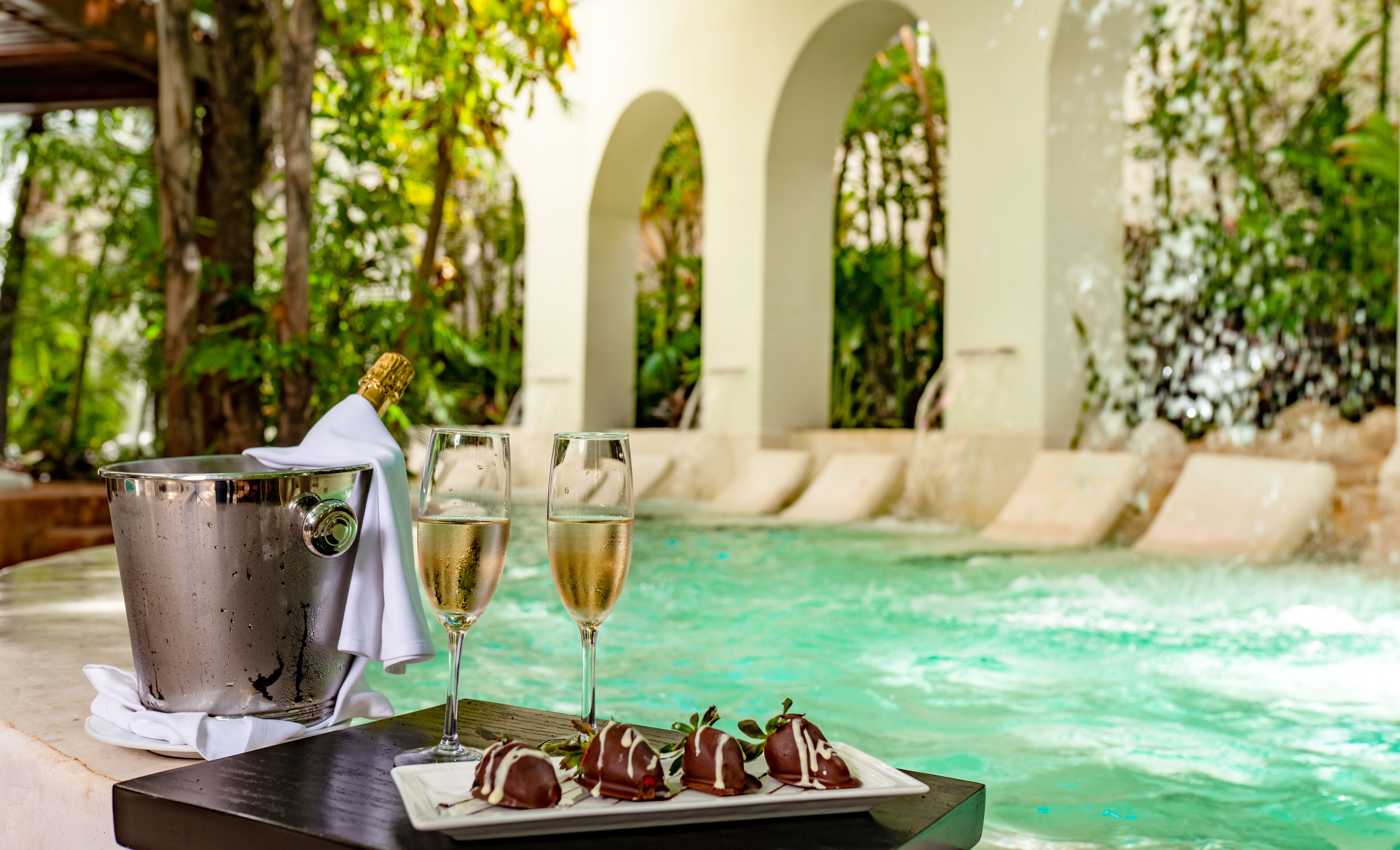 Cenário à beira da piscina com um balde de champanhe, duas taças de champanhe e morangos cobertos de chocolate em um prato.