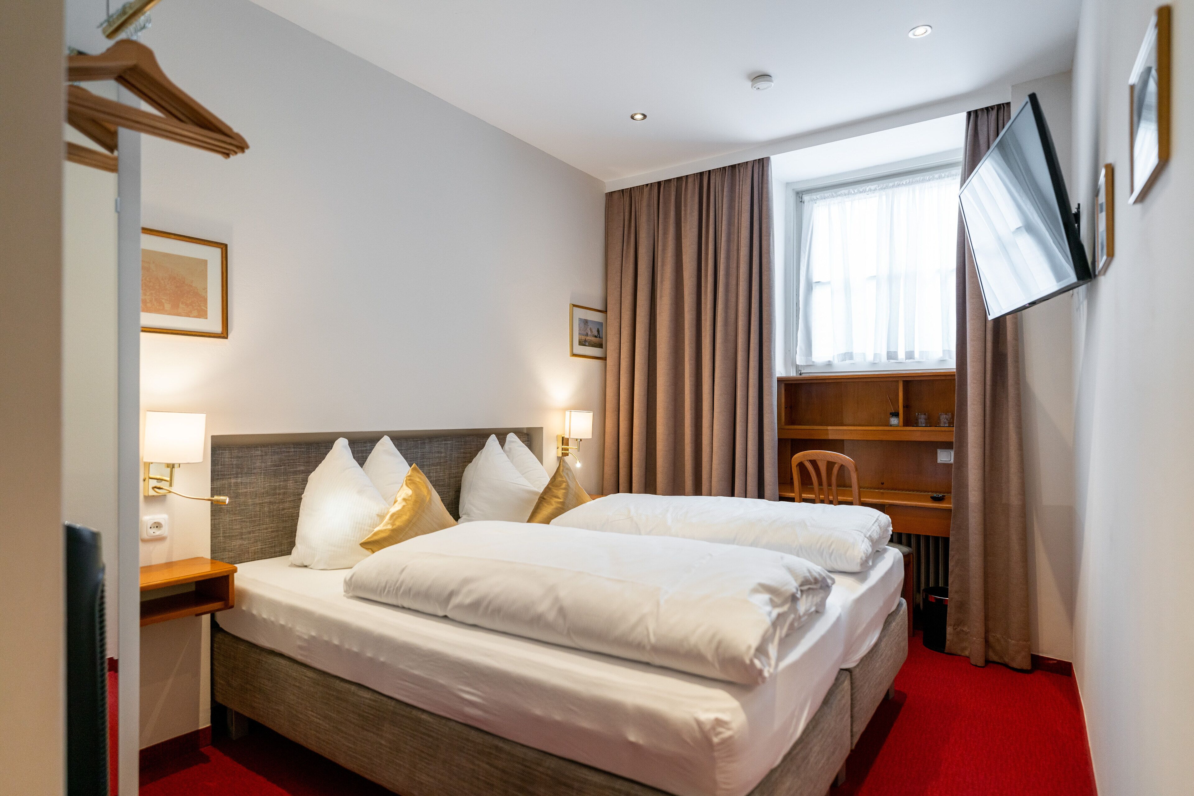 Quarto de hotel com cama de casal, mesas de cabeceira, escrivaninha, cadeira, televisão e janela com cortinas.