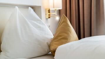 Cama bem arrumada com almofadas brancas e douradas, um abajur e uma cortina ao fundo.
