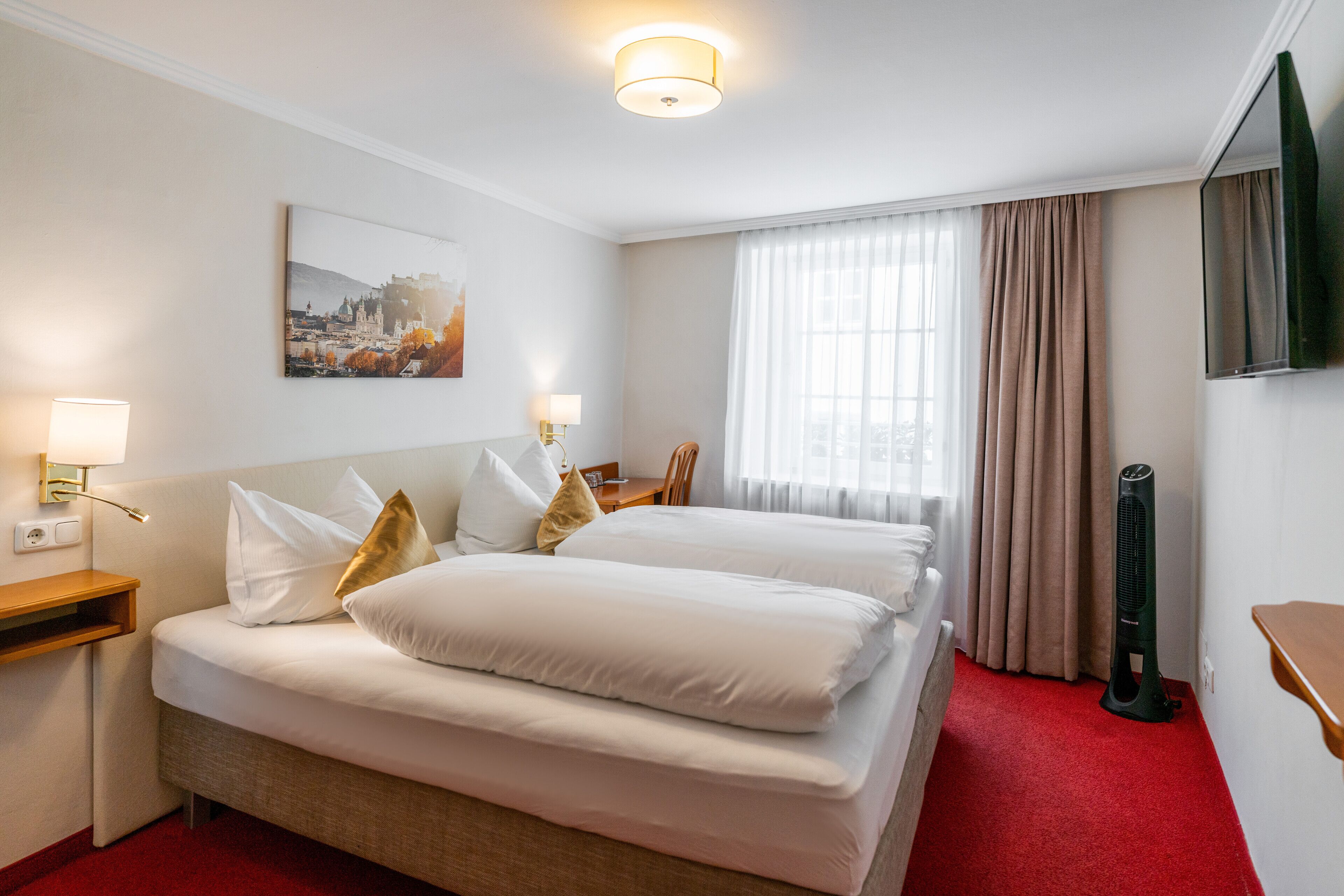 Quarto de hotel com uma cama grande, duas mesas de cabeceira com luminárias, uma TV de tela plana, uma janela ampla com cortinas e um quadro na parede.