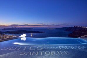 Piscina infinita com a inscrição 'Suites of the Gods Santorini', com vista para a paisagem costeira.
