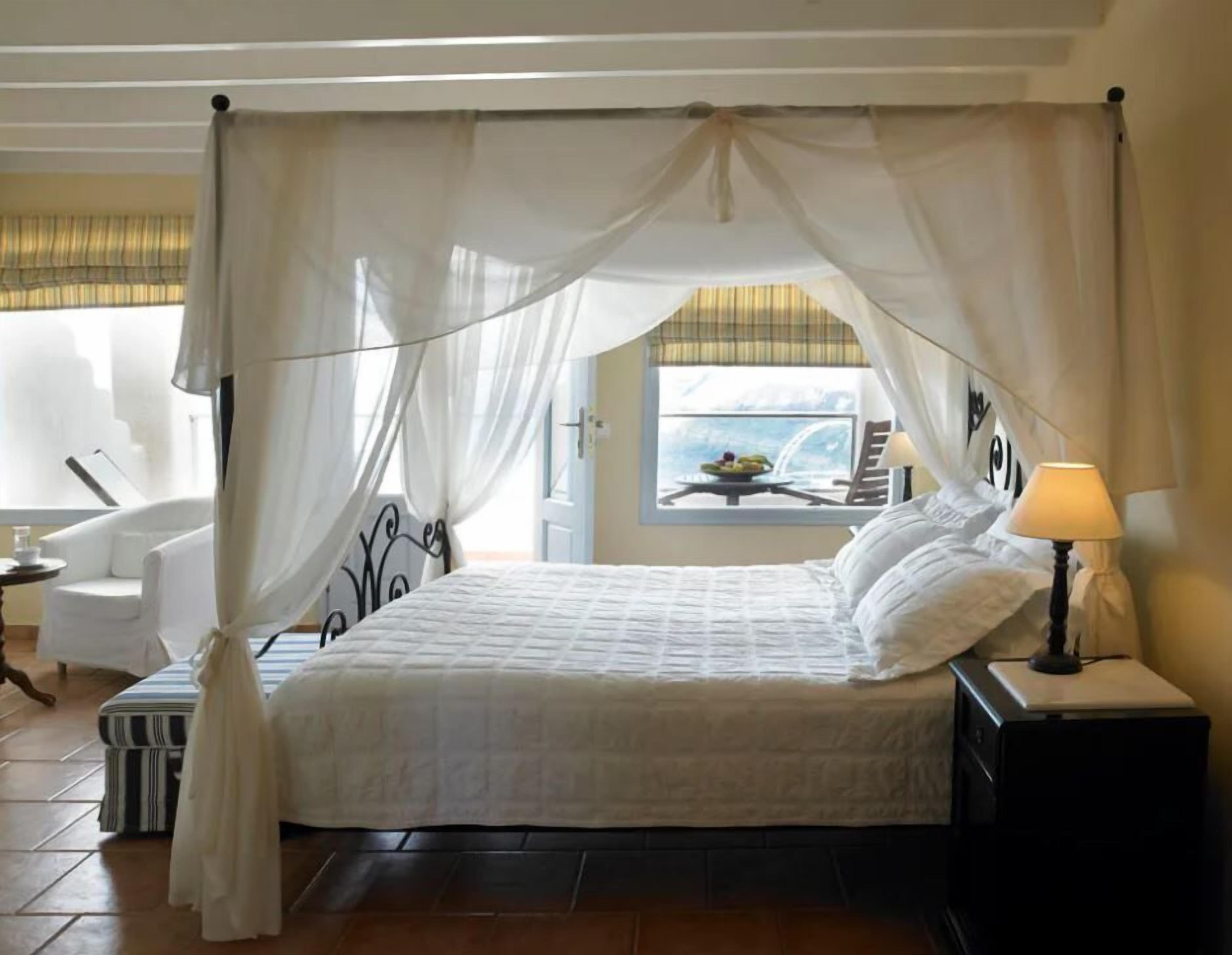Quarto com cama de dossel, mesa de cabeceira com abajur, uma cadeira e vista para o mar pela janela.