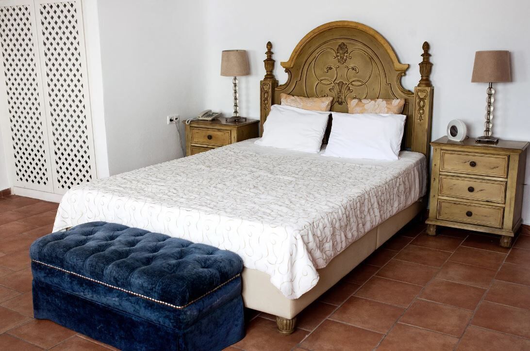 Uma cama com cabeceira de madeira, duas mesas de cabeceira, um criado-mudo, um banquinho estofado azul e um cobreleiro branco estampado.