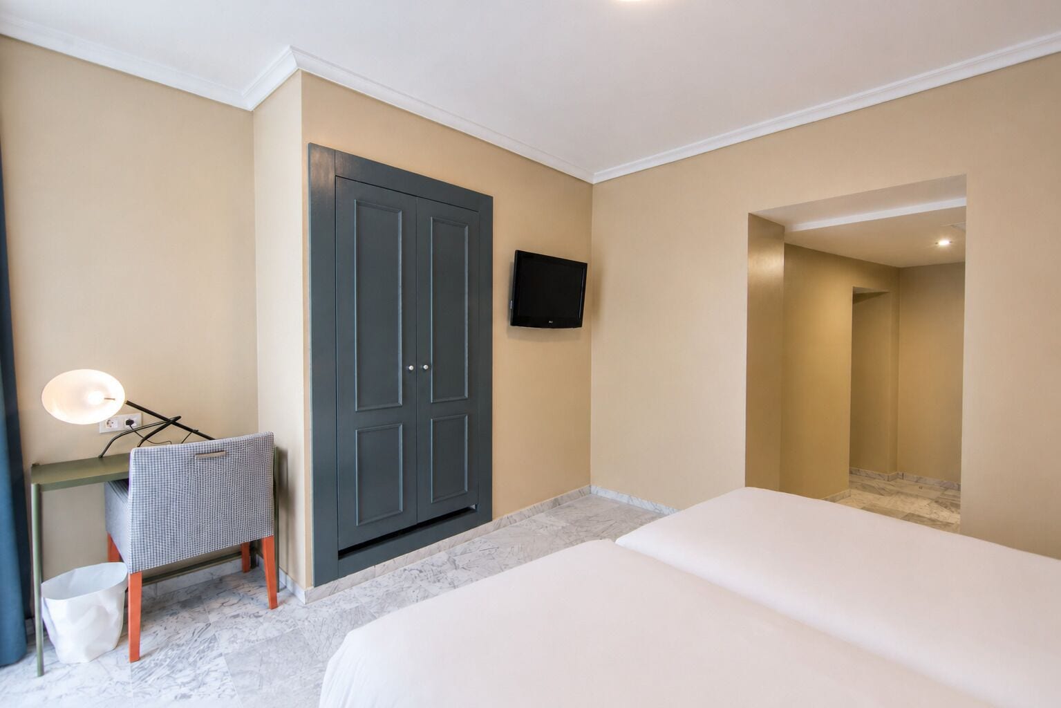 Quarto de hotel com cama, escrivaninha com abajur, armário e TV fixada na parede.