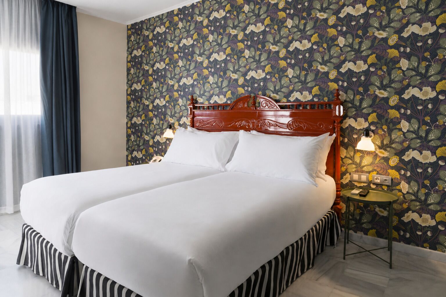 Cama de casal com cabeceira de madeira, roupa de cama branca, saia listrada e papel de parede floral.