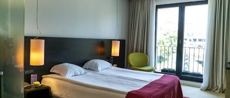 Quarto de hotel com uma cama grande, duas luminárias de cabeceira, uma cadeira verde e vista para o exterior por uma janela ampla.