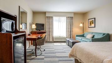 Quarto de hotel com uma cama, uma escrivaninha com cadeira, televisão, micro-ondas, abajur e janela com cortinas.