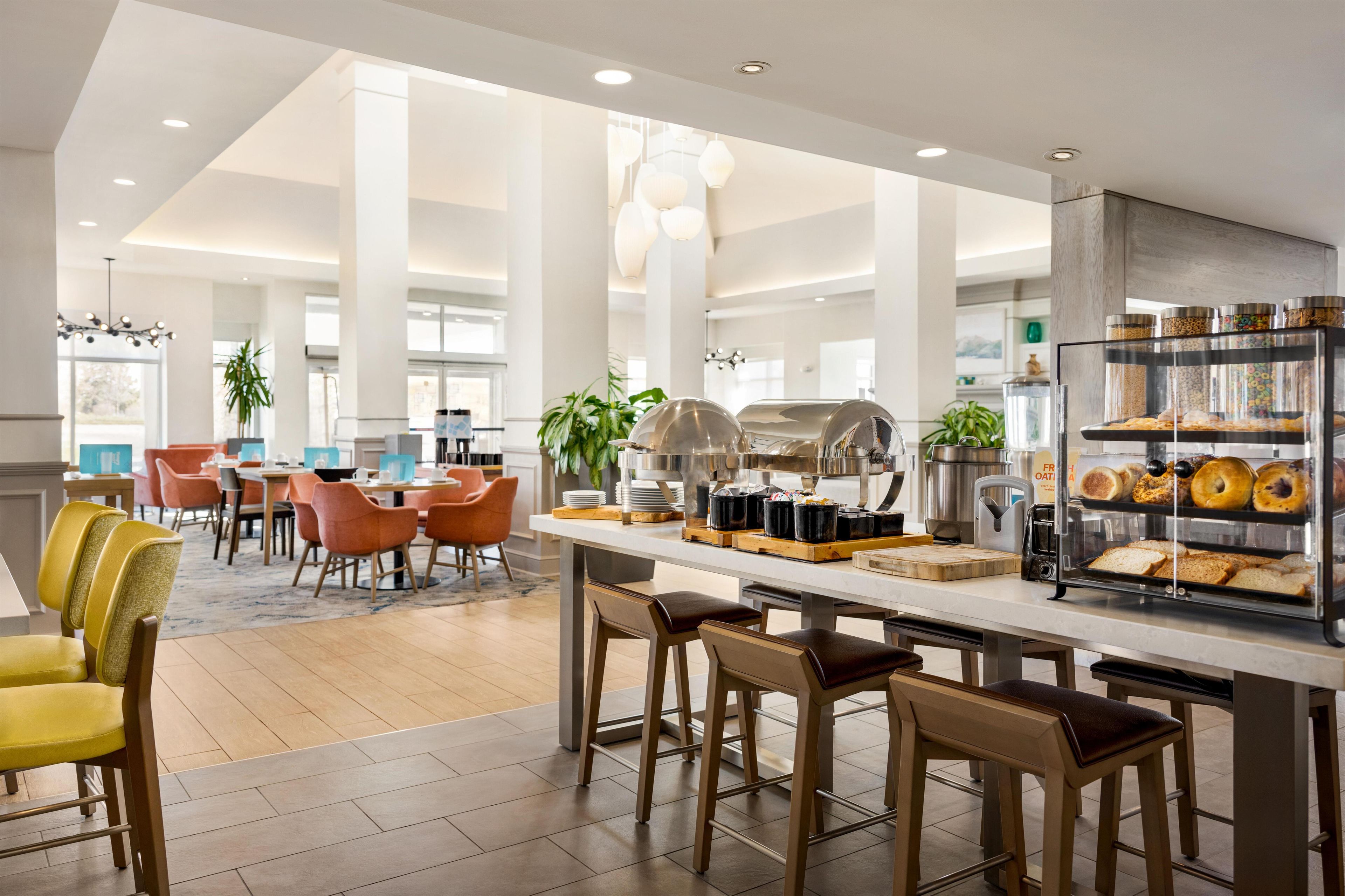 Espaço de refeições de hotel moderno, com pé direito alto, grandes janelas e uma ilha central com banquetas.