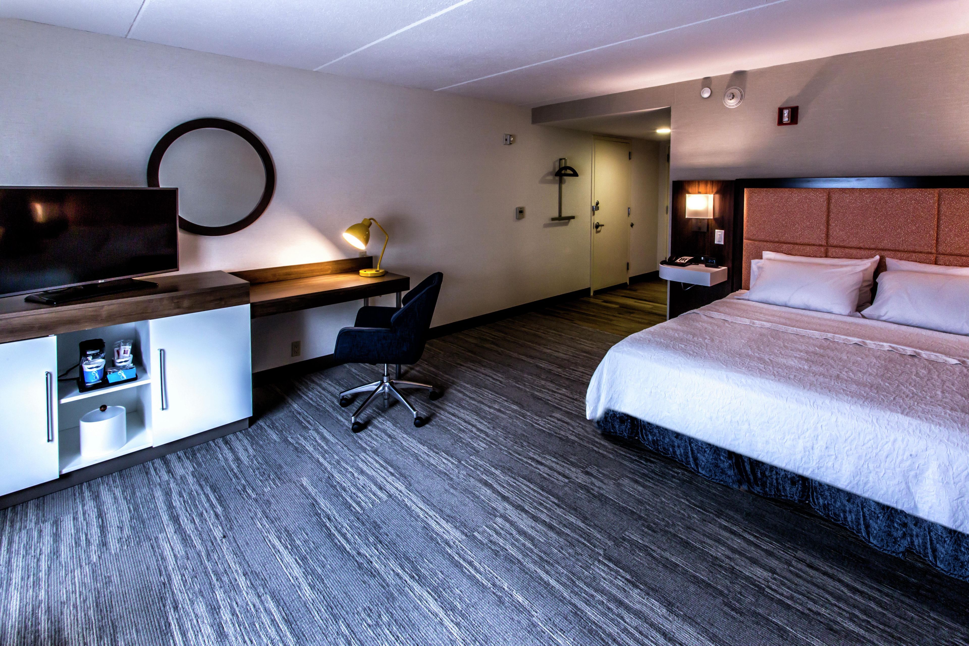 Quarto de hotel com cama, escrivaninha, cadeira, televisão e minibar.