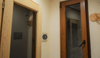 Um corredor com uma porta de madeira e uma porta de vidro, ambas dando para outro quarto. Há uma placa de aviso e um painel de controle na parede.