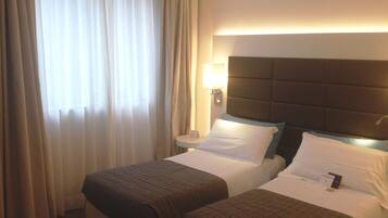 Quarto de hotel com duas camas, mesa de cabeceira, abajur e janela com cortinas.