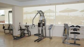 Uma academia com diversos aparelhos de exercícios, amplas janelas com vista para uma praia e guarda-sóis, e piso de madeira.