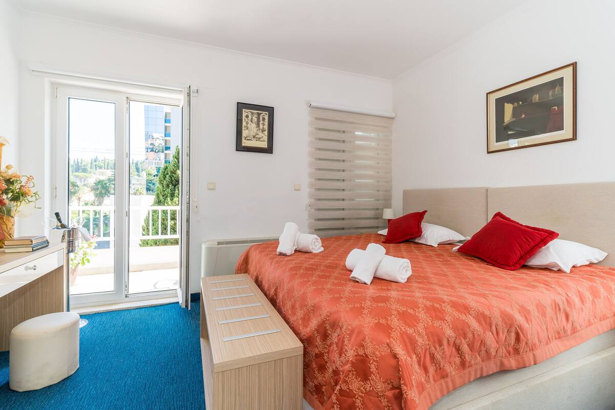 Quarto de hotel com cama, escrivaninha, cadeira, um banquinho pequeno, janela com persianas e vista para o exterior.