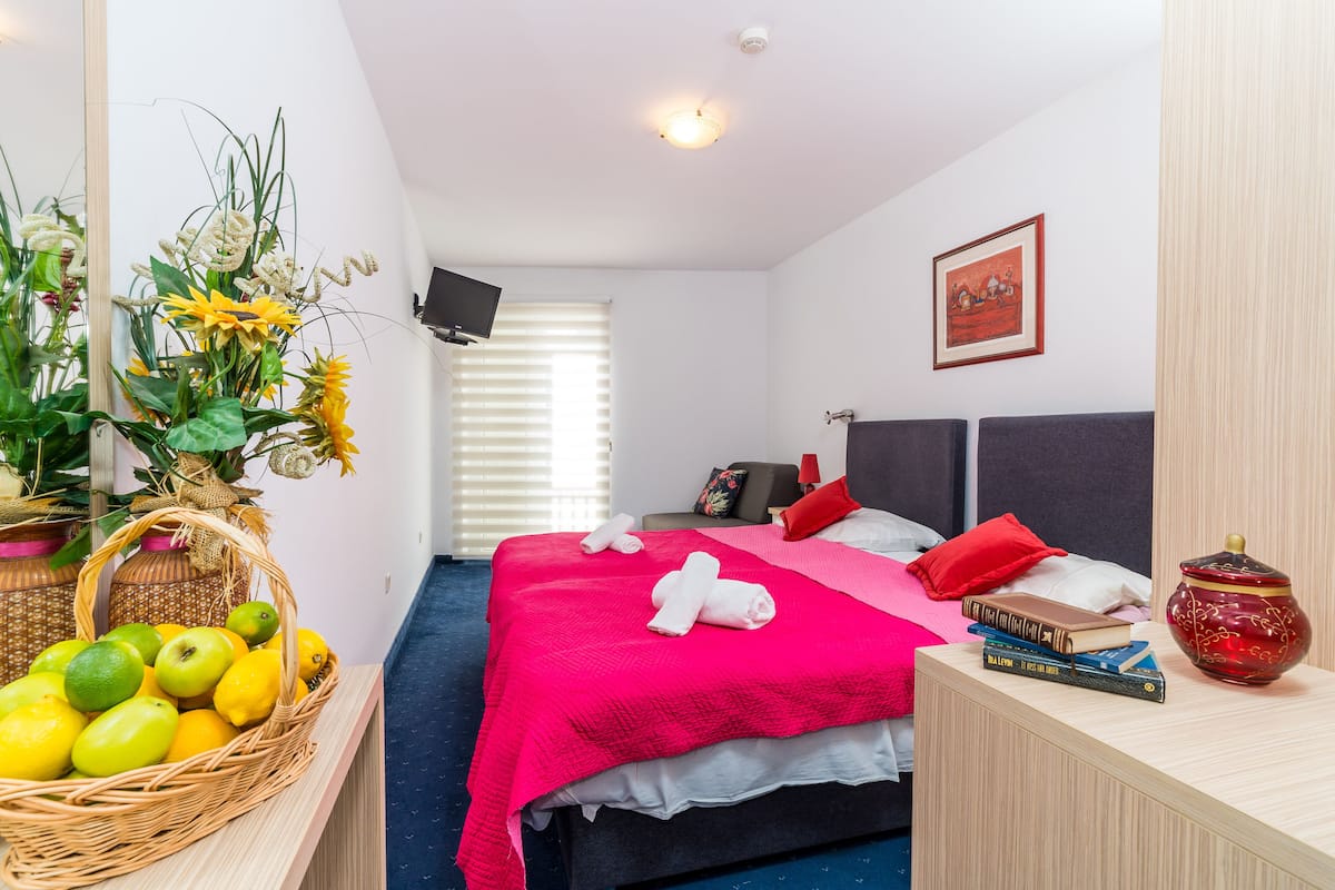 Quarto de hotel com cama, mesa de cabeceira com livros e um recipiente decorativo, cesta de vime com frutas e uma TV na parede.
