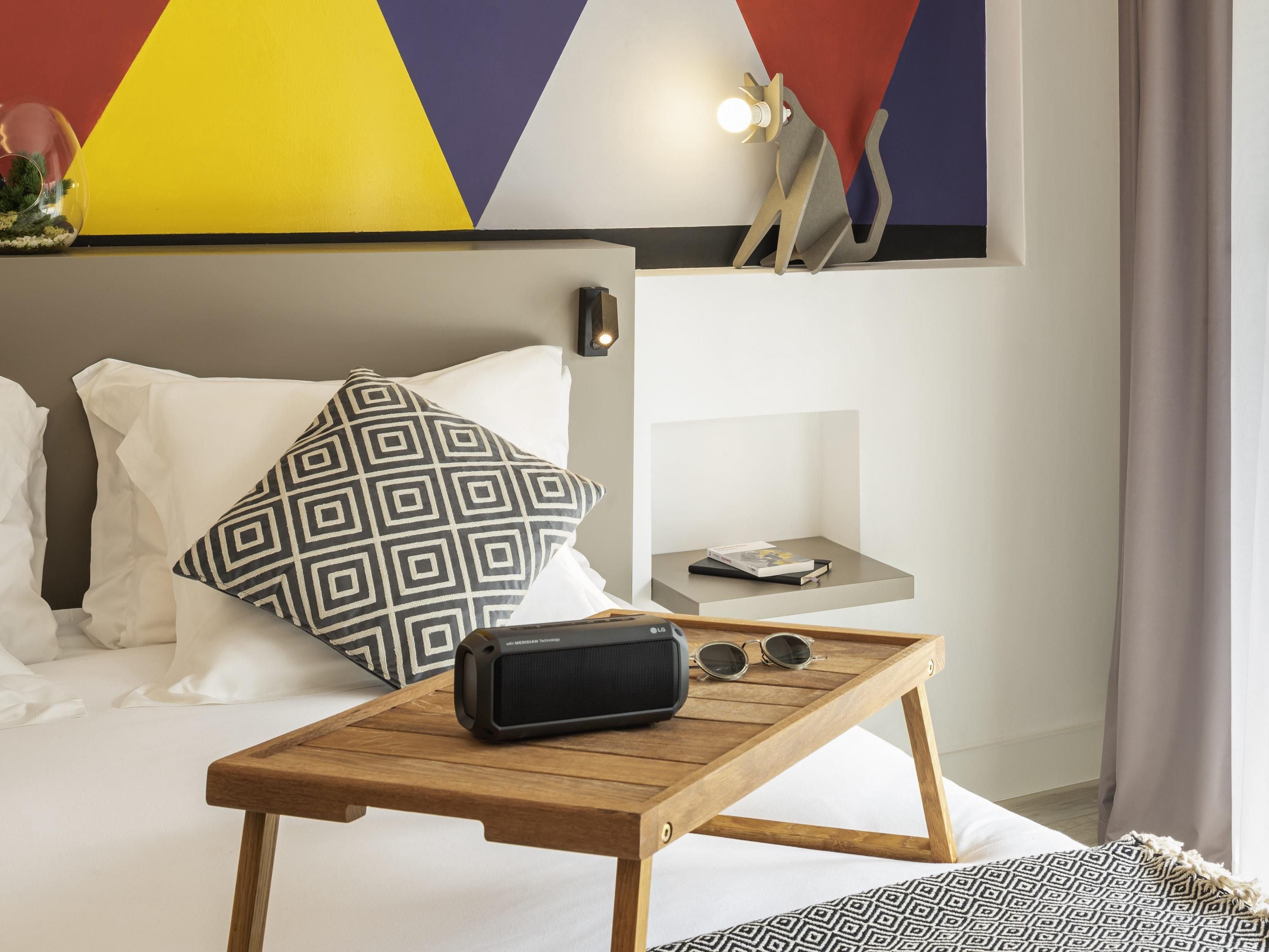 Quarto de hotel com uma cama, uma mesa de cabeceira com um alto-falante e óculos de sol, uma pequena estante com livros e uma decoração de parede colorida.