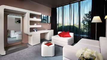 Quarto de hotel moderno com sofá branco, almofada vermelha, banqueta branca, escrivaninha com espelho e grandes janelas com vista para vegetação.