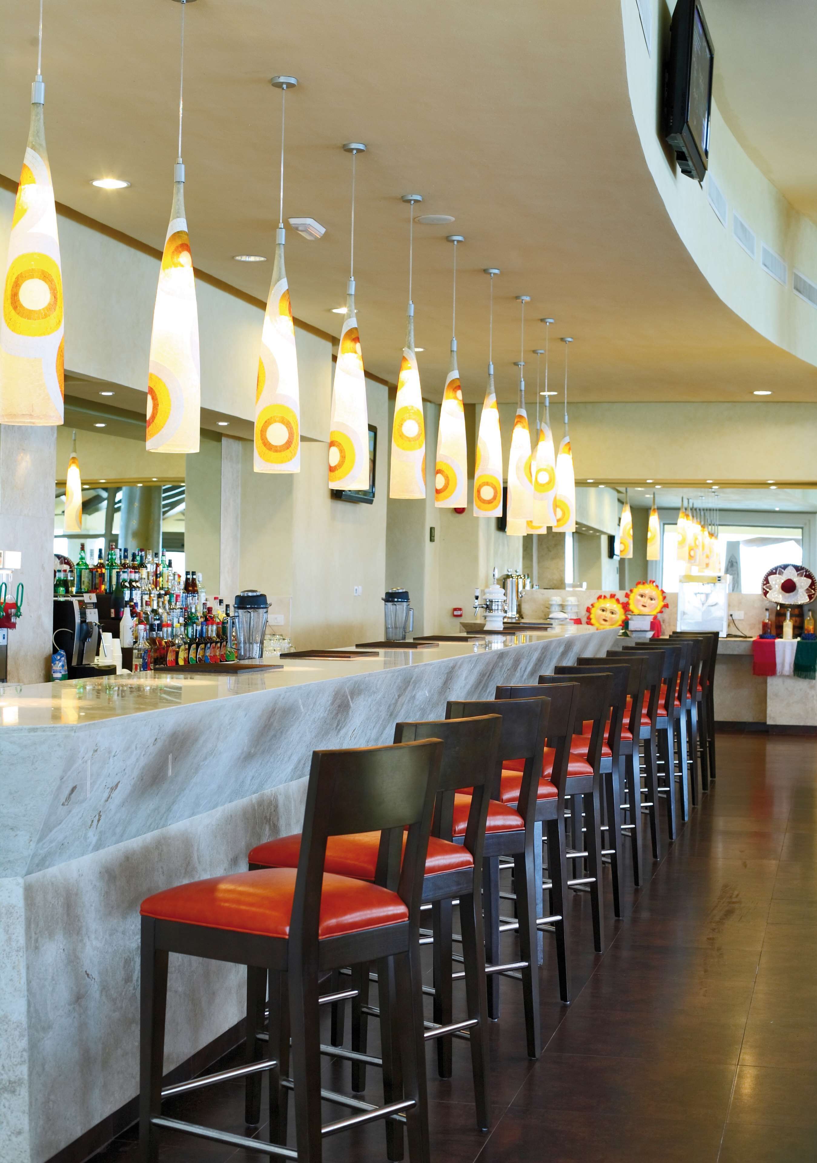 Uma área de bar com um balcão longo, várias luminárias pendentes e fileiras de banquetas.