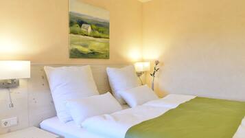 Quarto de hotel com uma cama, almofadas brancas, um cobertor verde, um quadro na parede e uma mesinha de cabeceira com uma luminária.