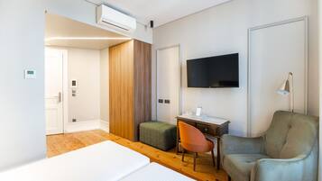 Quarto de hotel moderno com piso de madeira, uma cama, escrivaninha com cadeira, televisão e uma janela grande.