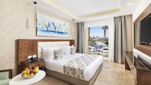 Quarto de hotel com uma cama grande, uma cesta de frutas, um quadro com veleiros, uma televisão e uma varanda com vista para palmeiras.