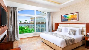 Quarto de hotel com uma cama grande, televisão, pintura e vista para áreas verdes e edifícios pela janela.