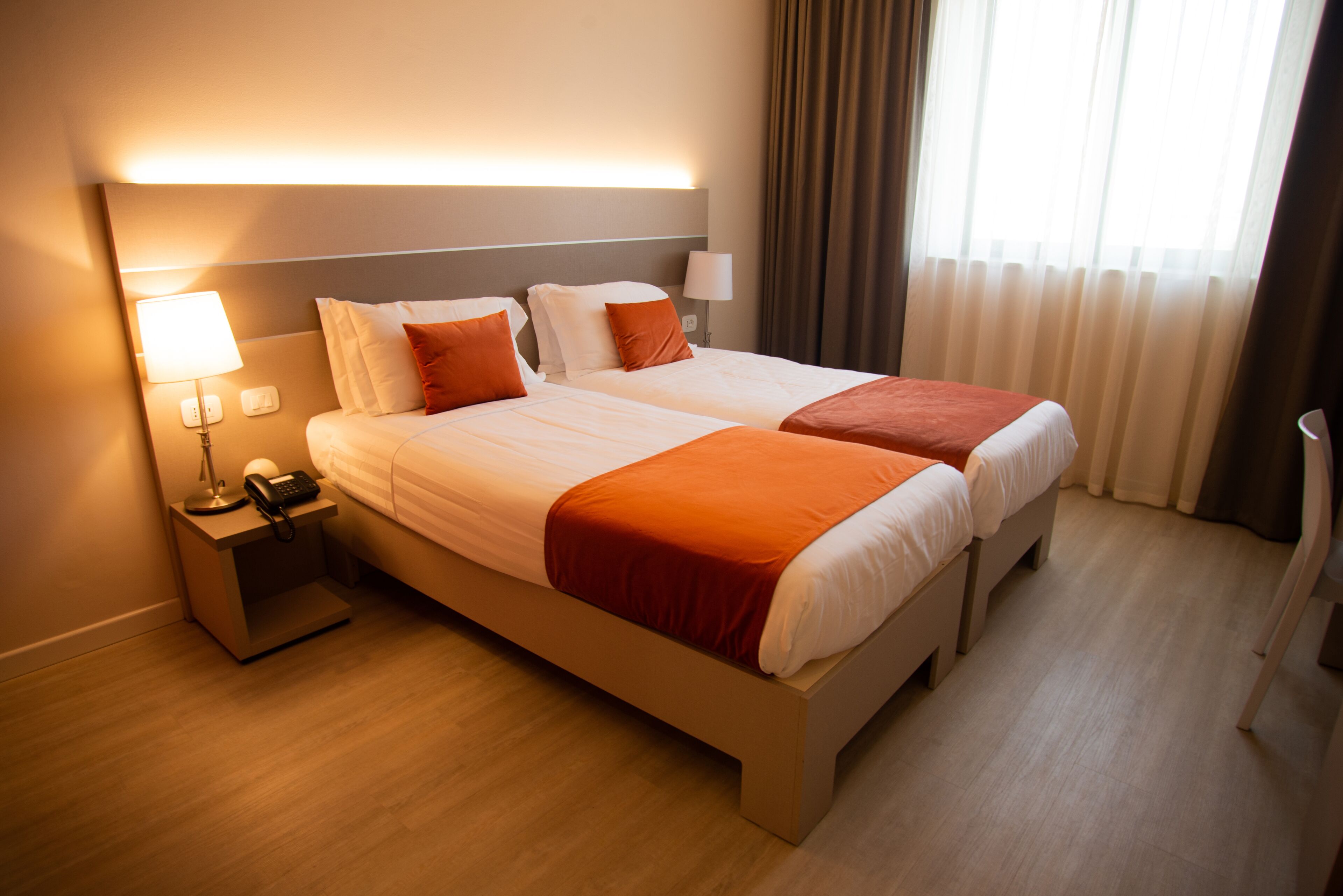 Uma cama de casal com cabeceira, duas mesas de cabeceira com luminárias e uma janela com cortinas.