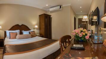 Quarto de hotel com uma cama grande, luminárias de cabeceira, uma escrivaninha com um vaso de flores e um aparelho de ar condicionado.
