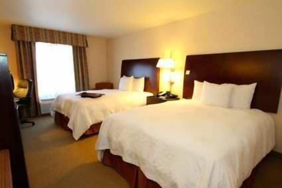Quarto de hotel com duas camas, uma mesa, uma cadeira e uma janela com cortinas.