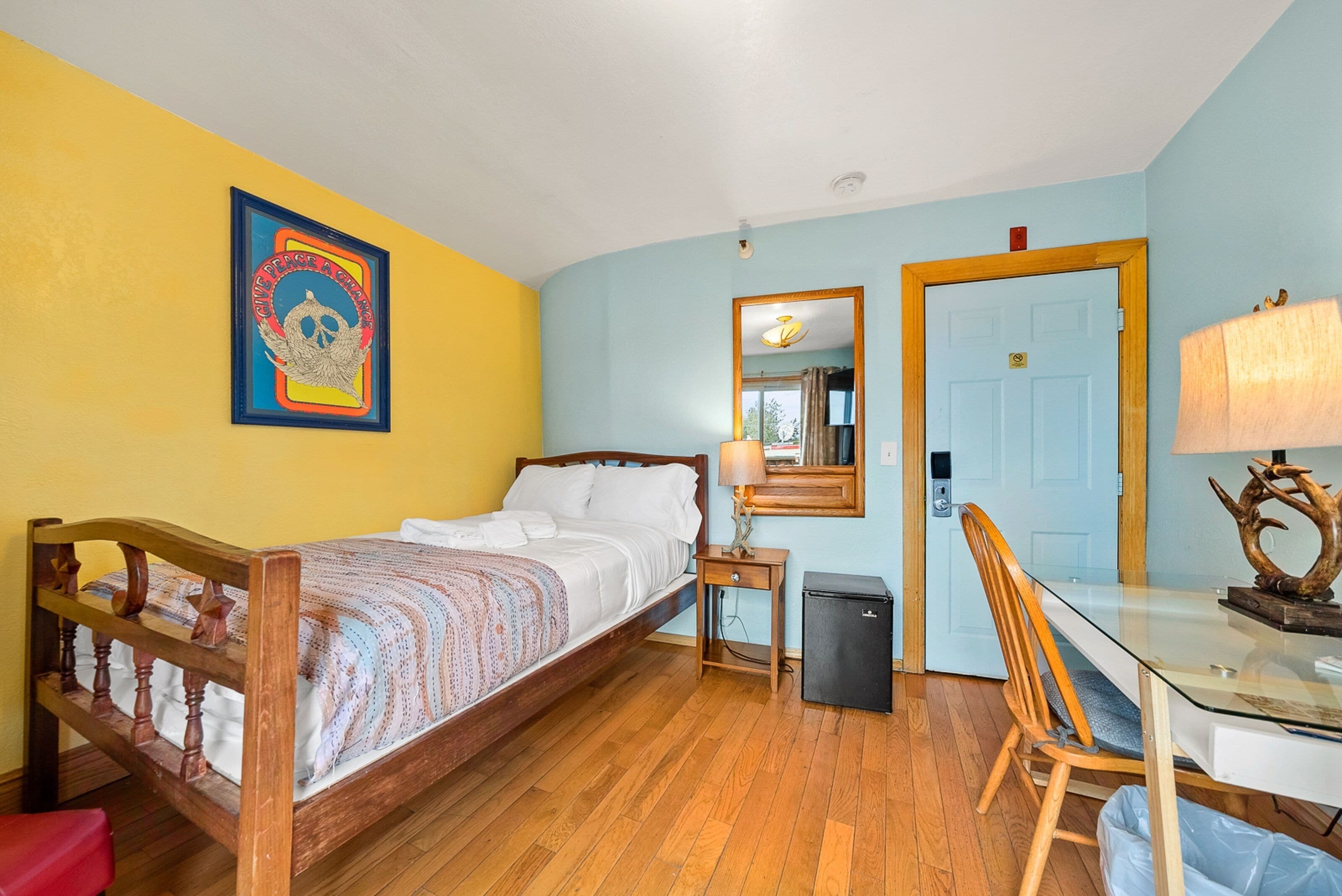 Quarto com cama de madeira, uma escrivaninha pequena com cadeira, um espelho e uma porta.